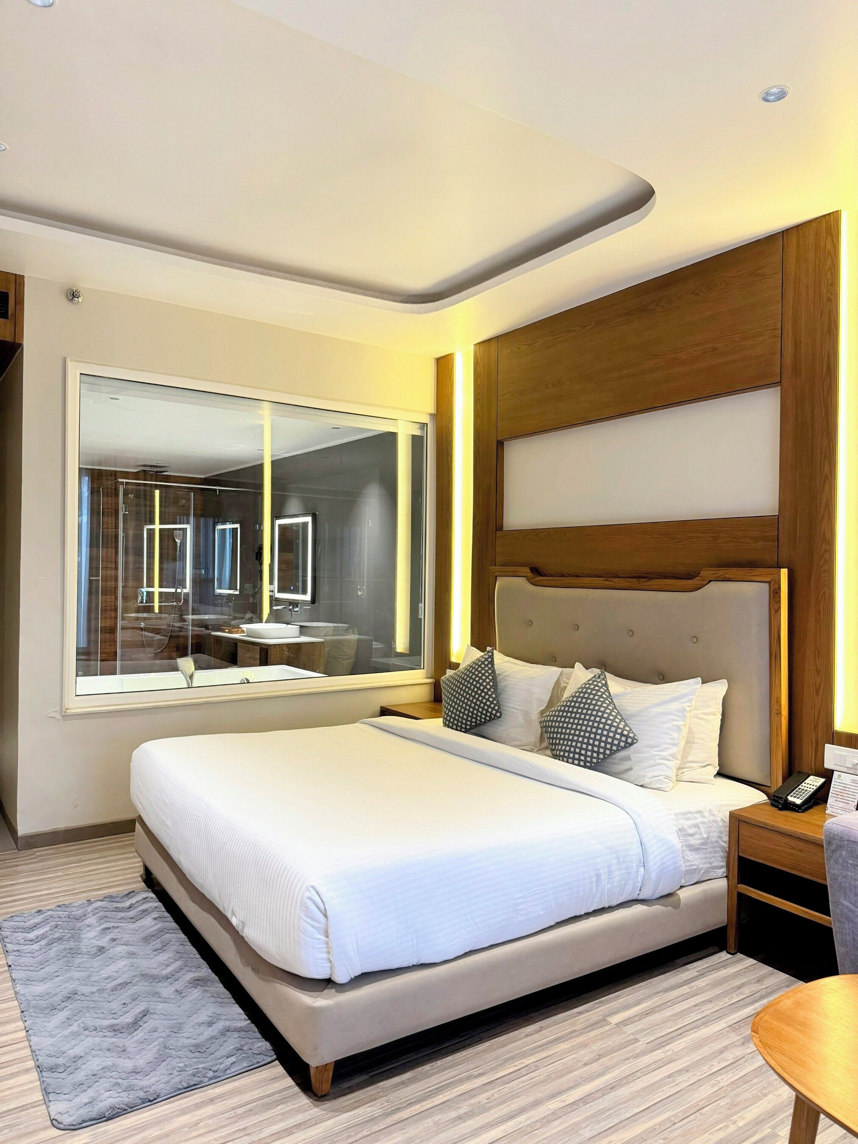 Quarto de hotel moderno com uma cama grande, mesa de cabeceira, espelho e vista para o banheiro.