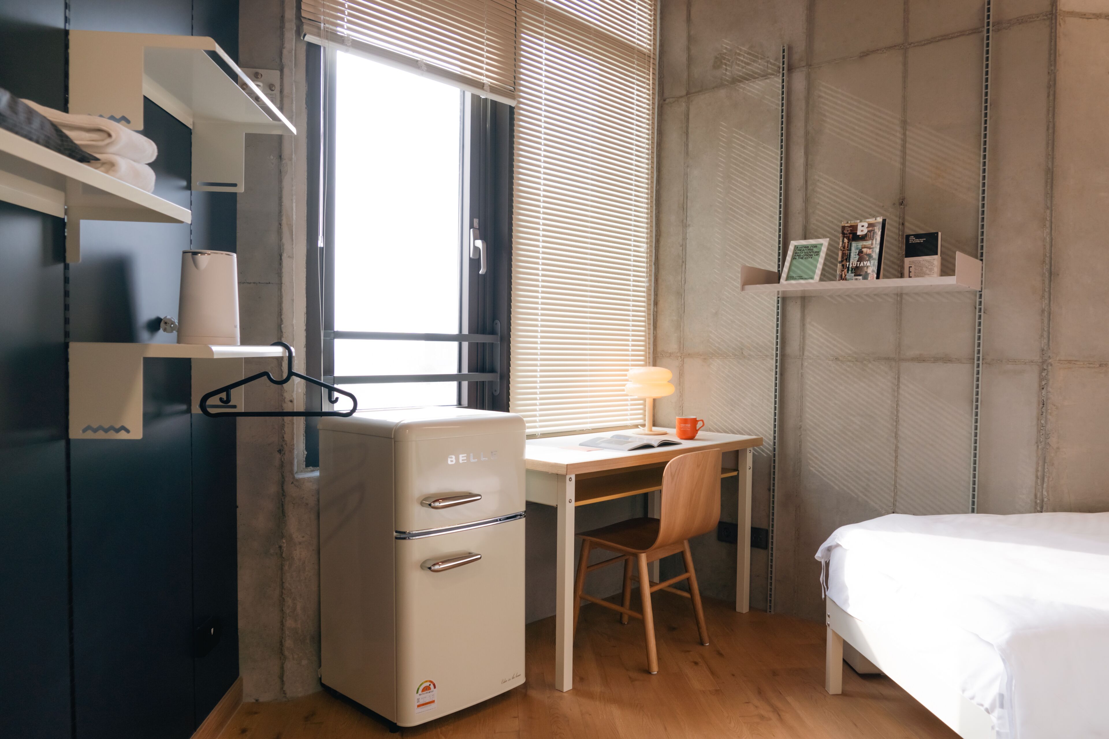 Quarto de hotel compacto com cama, escrivaninha com cadeira, pequeno espaço de cozinha e janela com persianas.