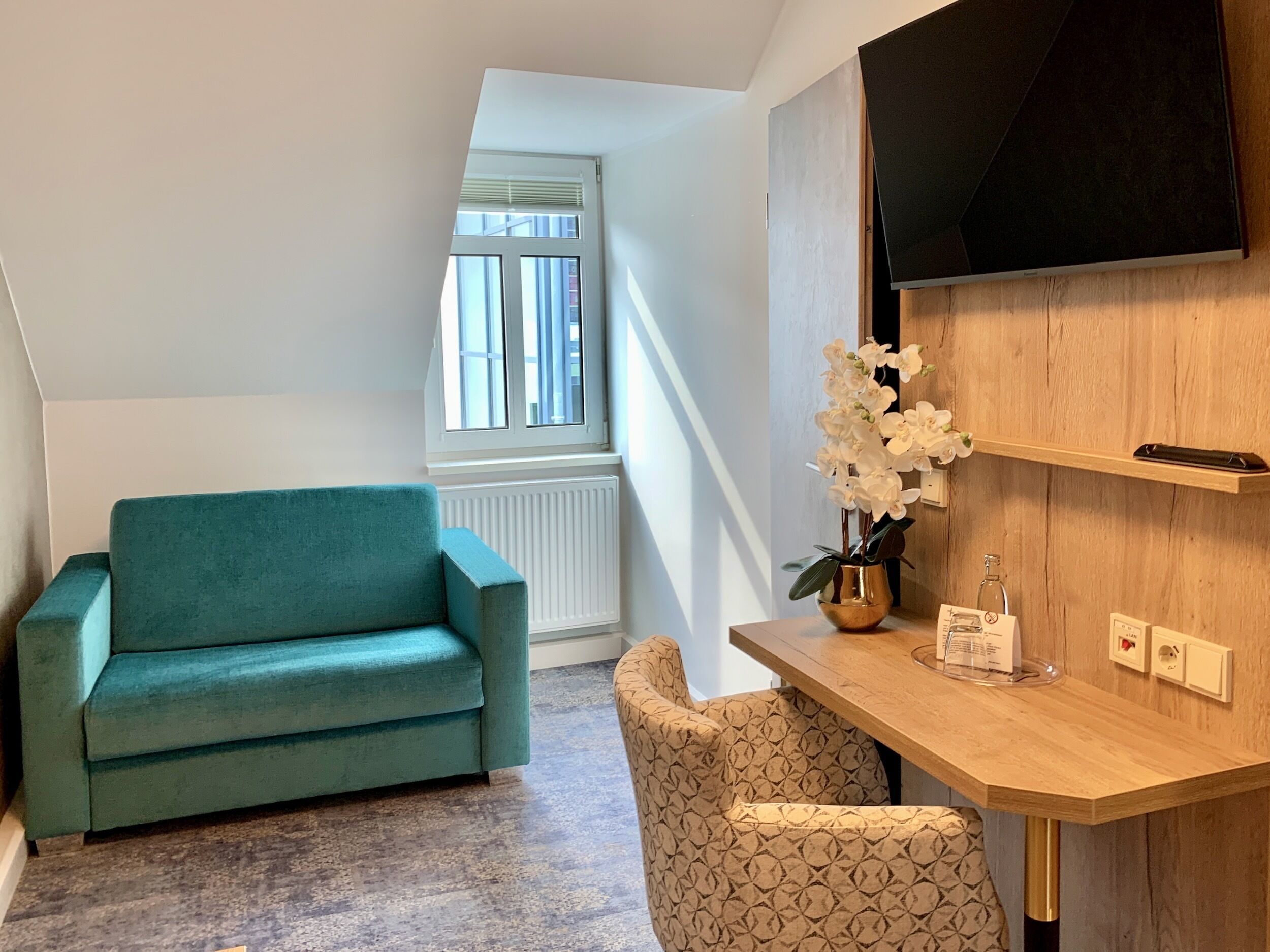 Quarto moderno com sofá verde-água, poltrona estampada, escrivaninha de madeira com televisão de tela plana e janela com persianas.