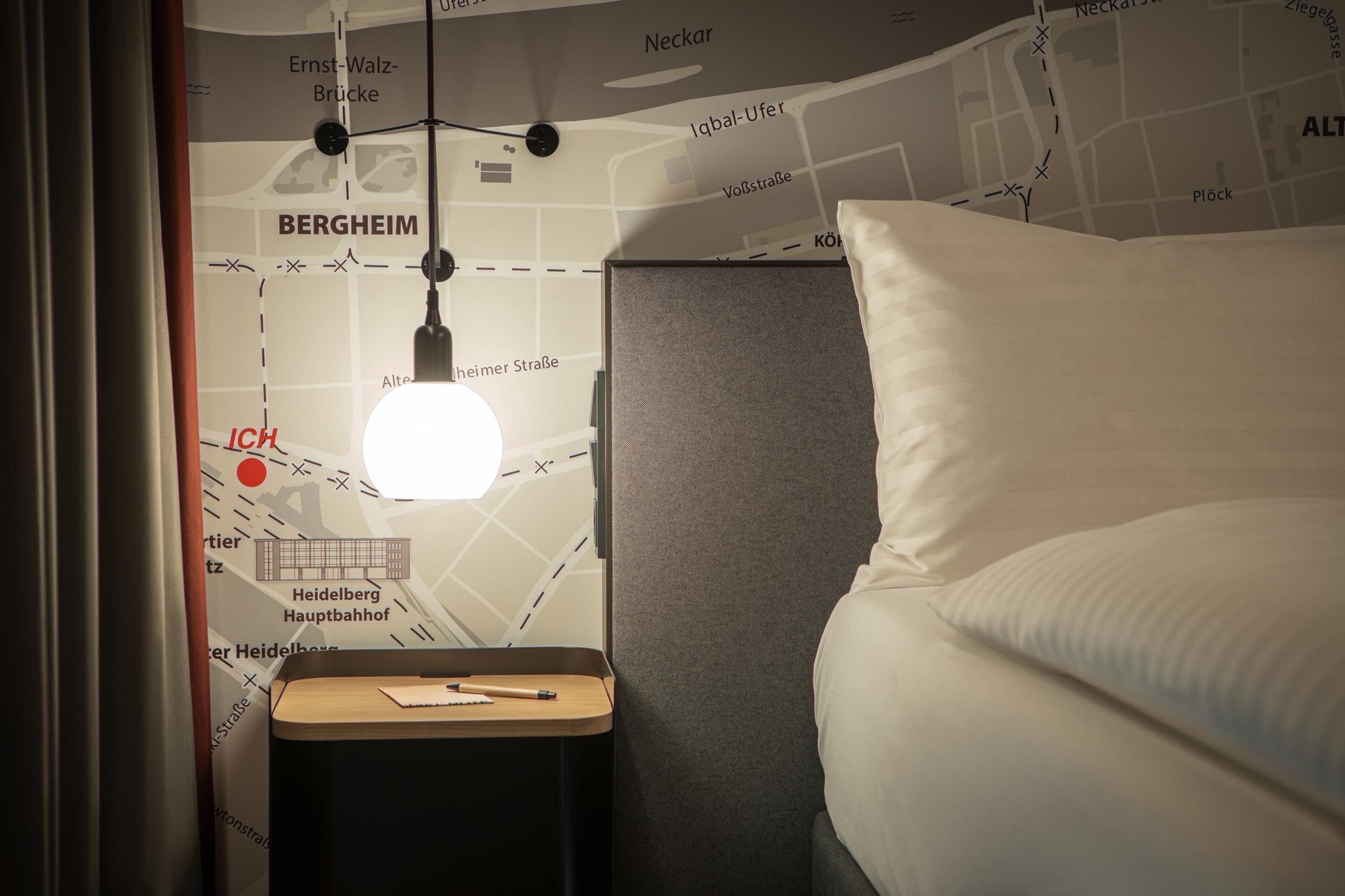 Quarto de hotel com parede de mapa, uma cama, mesa de cabeceira com abajur e uma mesinha de madeira.