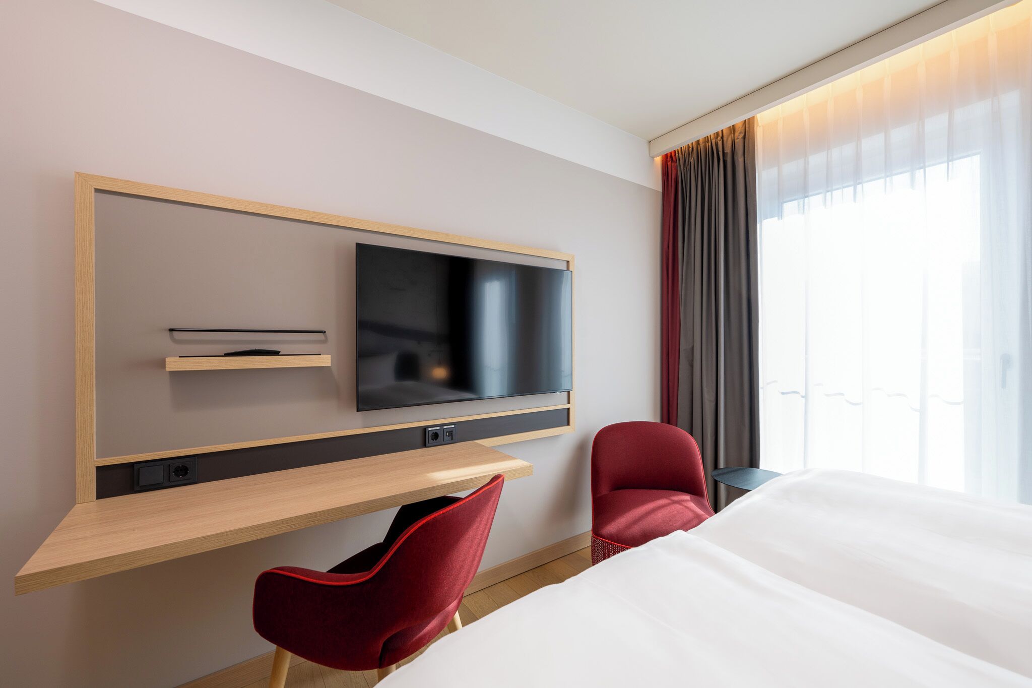 Quarto de hotel moderno com uma cama grande, uma televisão presa a uma prateleira de madeira, uma mesa de trabalho com uma cadeira e uma janela com cortinas.