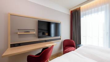 Quarto de hotel moderno com uma cama grande, uma televisão presa a uma prateleira de madeira, uma mesa de trabalho com uma cadeira e uma janela com cortinas.