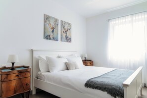 Quarto com cama branca, mesa de cabeceira de madeira, luminária e duas pinturas penduradas na parede.