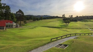 Um campo de golfe com um edifício de telhado vermelho, um caminho sinuoso e uma vista panorâmica de colinas e árvores.