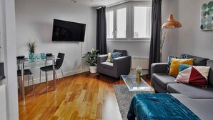 Uma sala de estar moderna com área de jantar, um sofá cinza, uma mesa de centro e uma TV de tela plana fixada na parede.