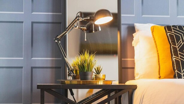 Quarto bem organizado com uma cama, uma mesa de cabeceira com abajur e um pequeno vaso de planta.