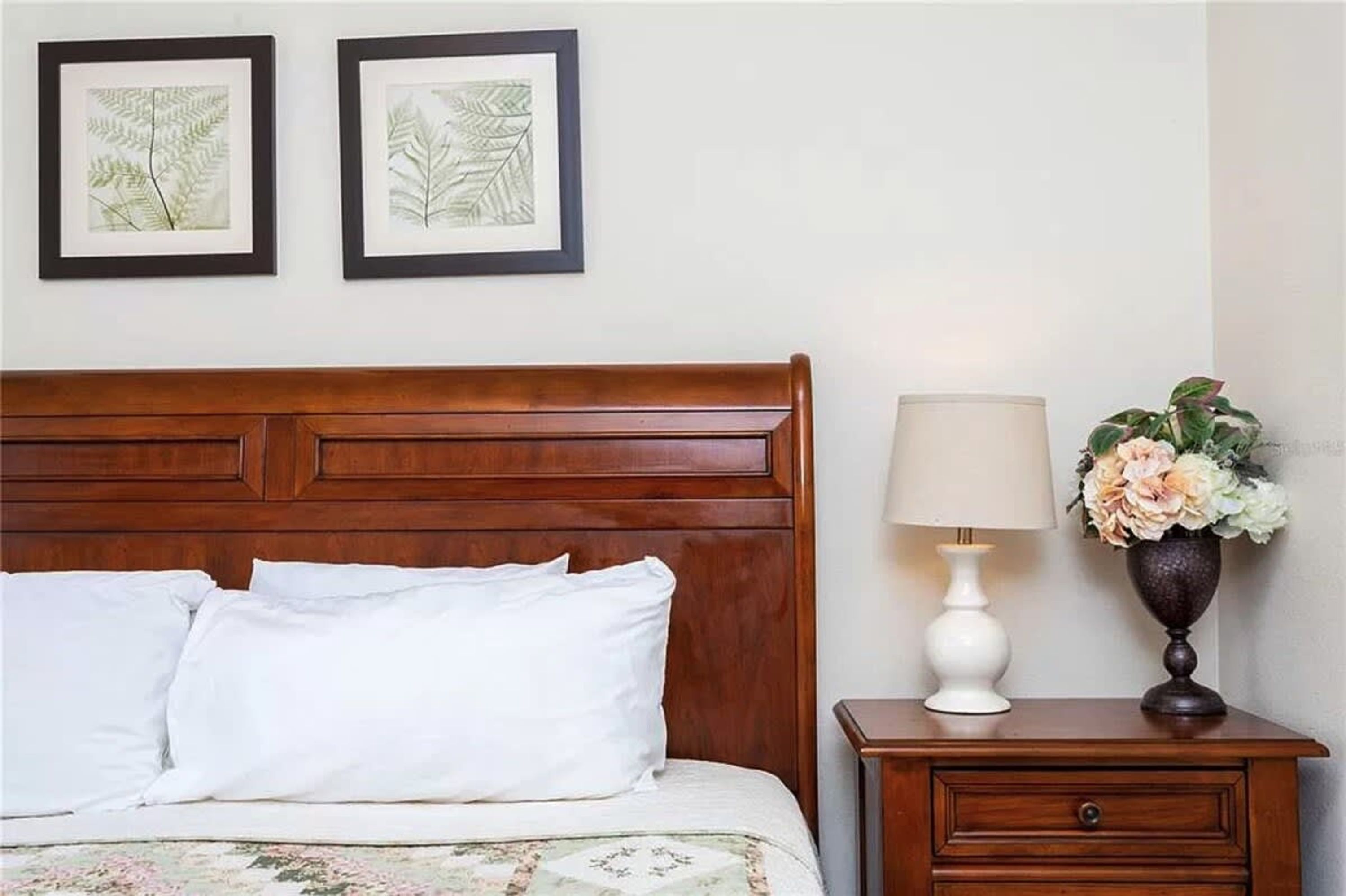Cama bem arrumada com lençóis brancos, cabeceira de madeira, mesa de cabeceira com abajur e um vaso de flores, e duas gravuras botânicas emolduradas na parede.
