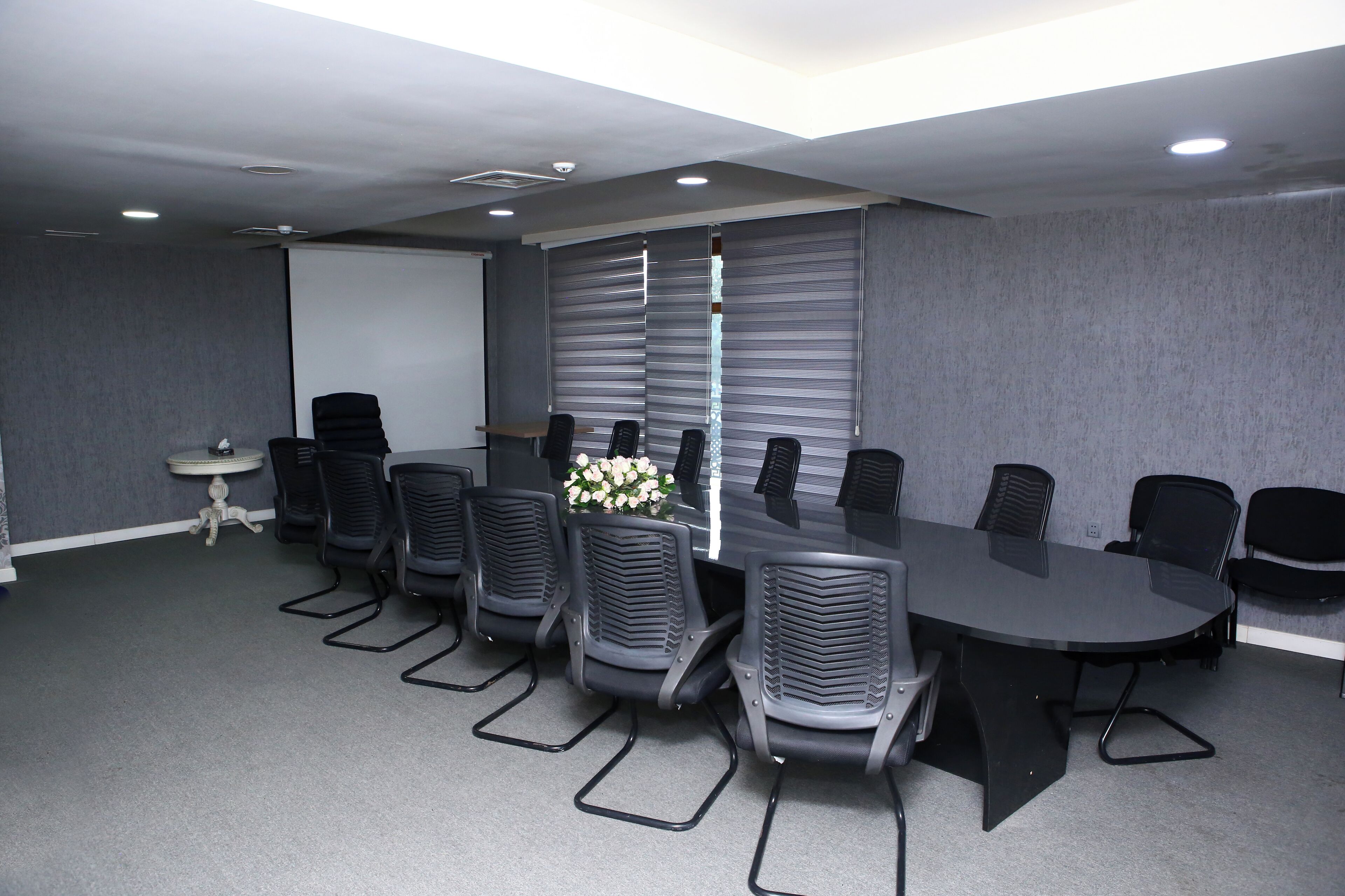 Uma sala de conferências com uma mesa grande, cadeiras pretas, uma tela de projetor e uma pequena mesa com um objeto decorativo.