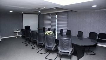 Uma sala de conferências com uma mesa grande, cadeiras pretas, uma tela de projetor e uma pequena mesa com um objeto decorativo.