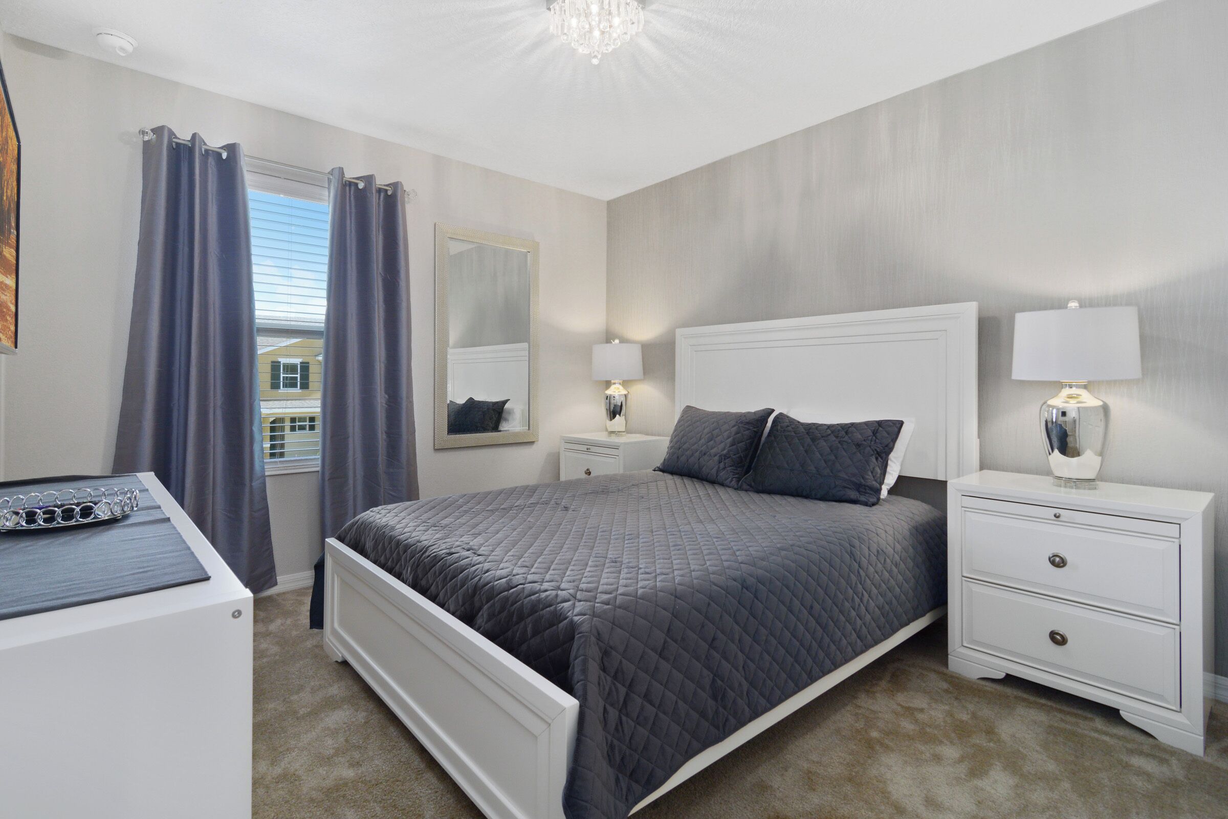 Quarto com cama, mesas de cabeceira, cômoda e janela com cortinas.