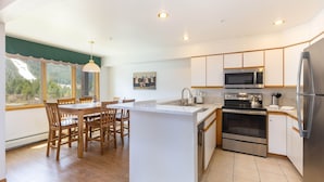 Cozinha moderna com armários de madeira, micro-ondas embutido, eletrodomésticos de aço inoxidável e um espaço de jantar com mesa e cadeiras.