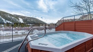 Uma banheira de hidromassagem com uma escada de metal, rodeada por um deck de madeira e uma cerca de metal, com vista para uma paisagem nevada e montanhas.