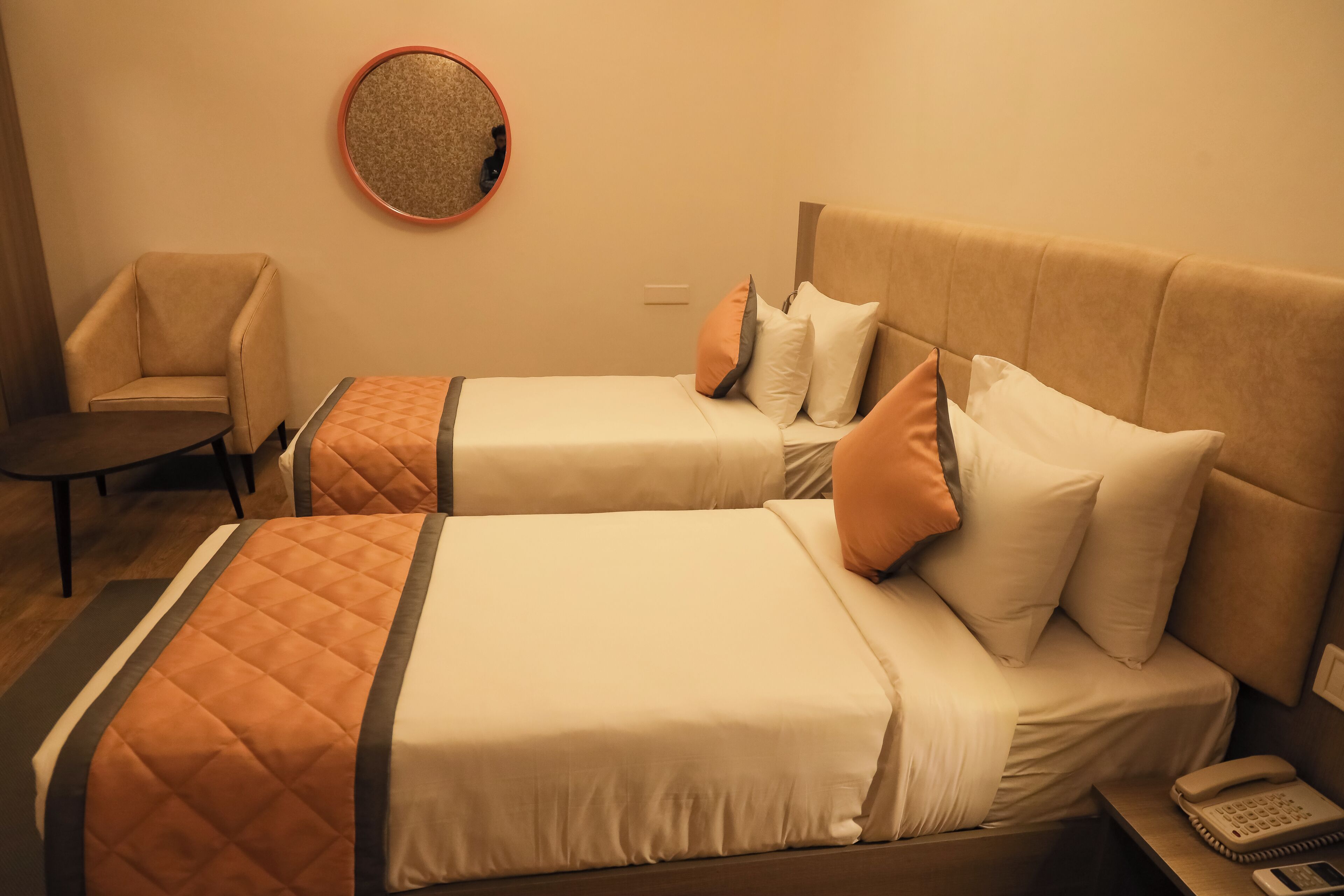 Quarto de hotel com duas camas, uma cadeira, um espelho redondo e um telefone ao lado da cama.