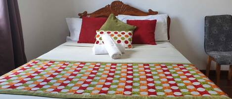 Uma cama bem arrumada com um espalha cama colorido estampado em bolinhas, uma cabeceira de madeira e uma cadeira cinza ao lado.
