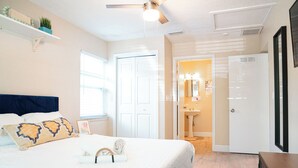 Quarto com cama, ventoinha no teto, porta branca e banheiro visível por uma porta aberta.