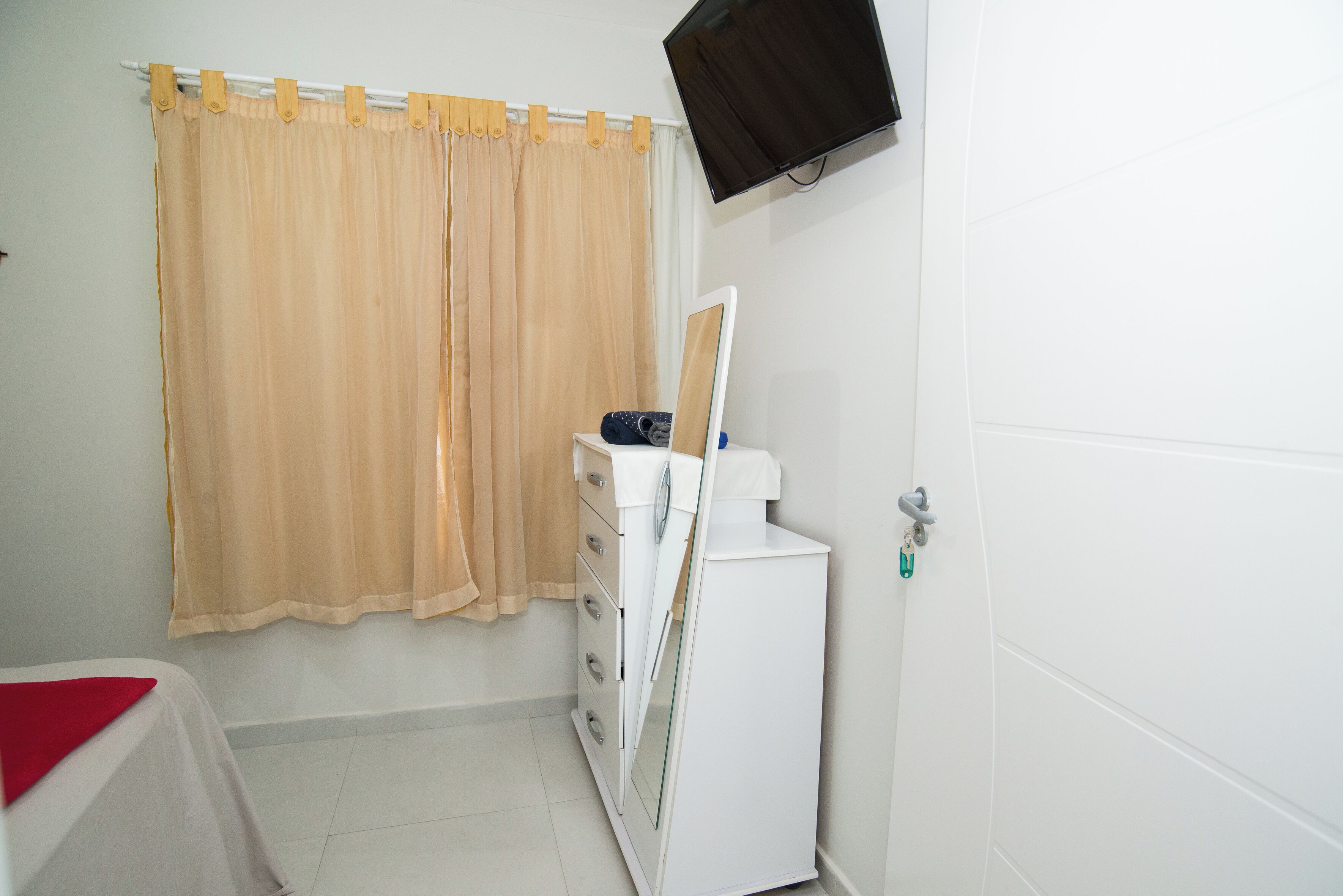 Quarto pequeno e limpo, com uma cama, cômoda, espelho e televisão na parede.
