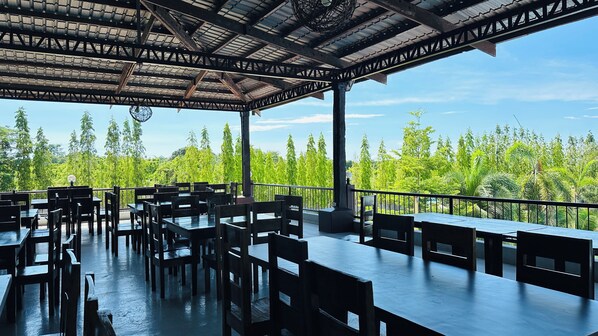 Uma área de refeições ao ar livre com mesas e cadeiras, cercada por uma grade de metal, com vista para uma paisagem verde exuberante.