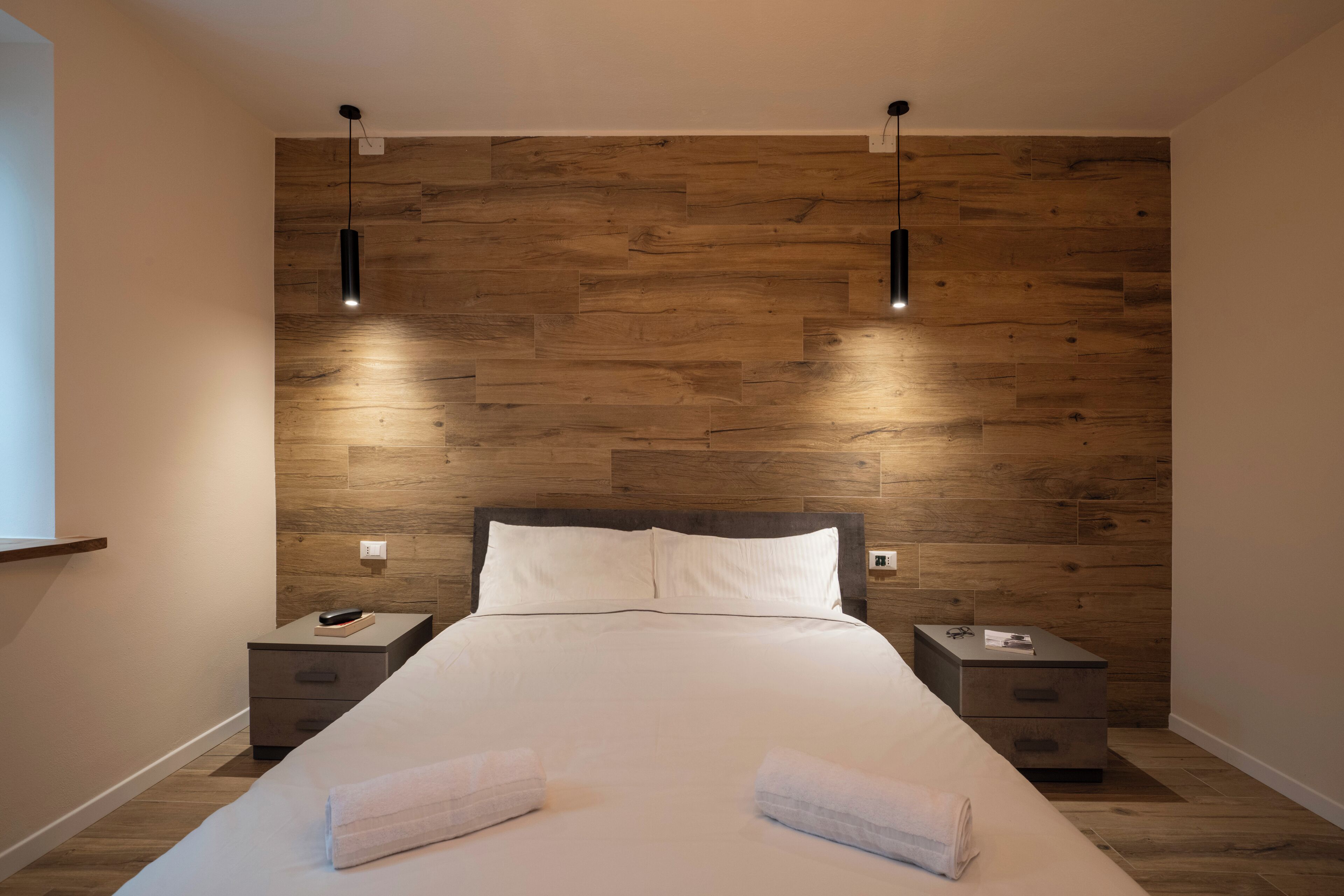 Quarto moderno com cabeceira de madeira, uma cama grande, duas mesas de cabeceira e duas luminárias pendentes.