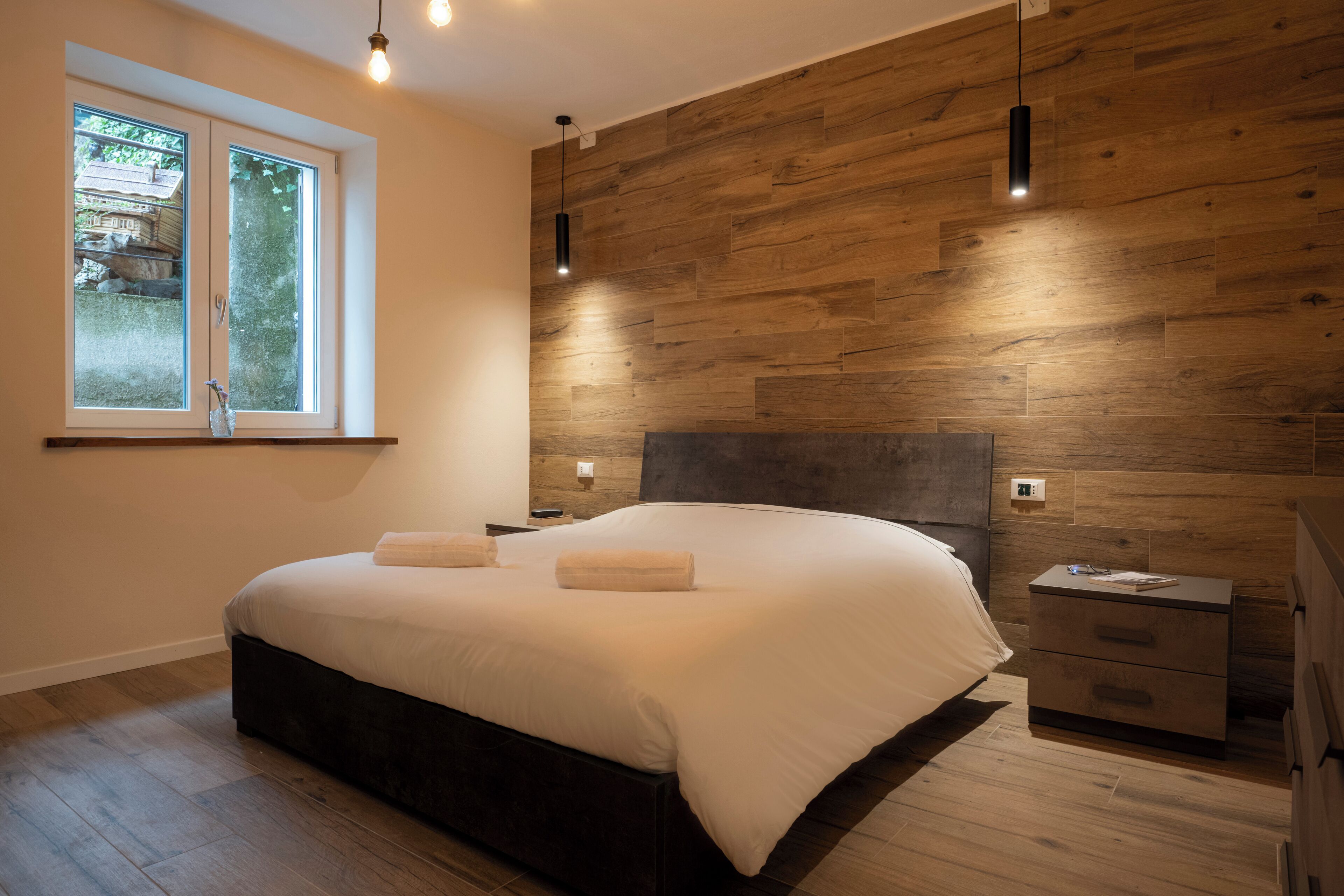 Quarto moderno com uma cama grande, painéis de parede de madeira, uma janela com vista e abajures de cabeceira.