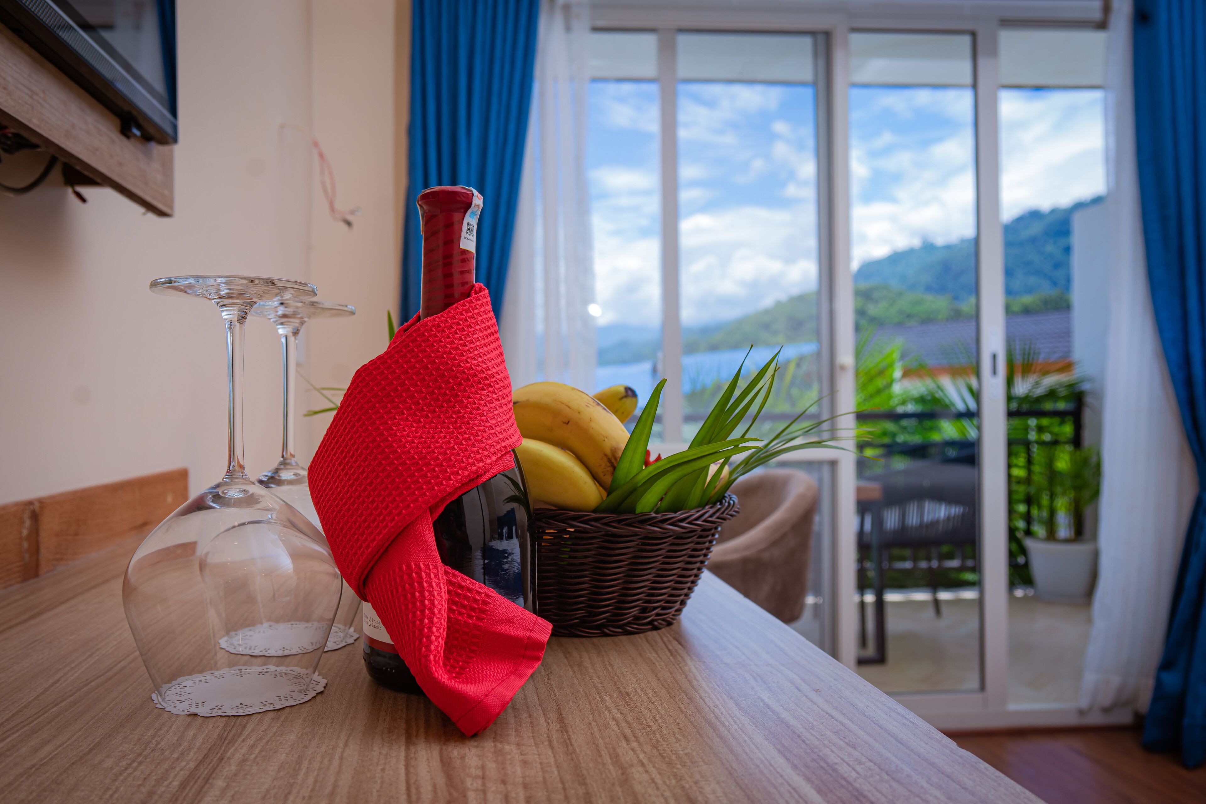 Uma mesa com uma garrafa de vinho, duas taças de vinho, um tecido vermelho e uma cesta de bananas e plantas verdes.