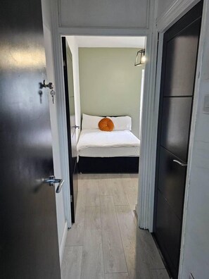 Um corredor estreito que dá para um quarto com uma cama, uma porta e um luminário.