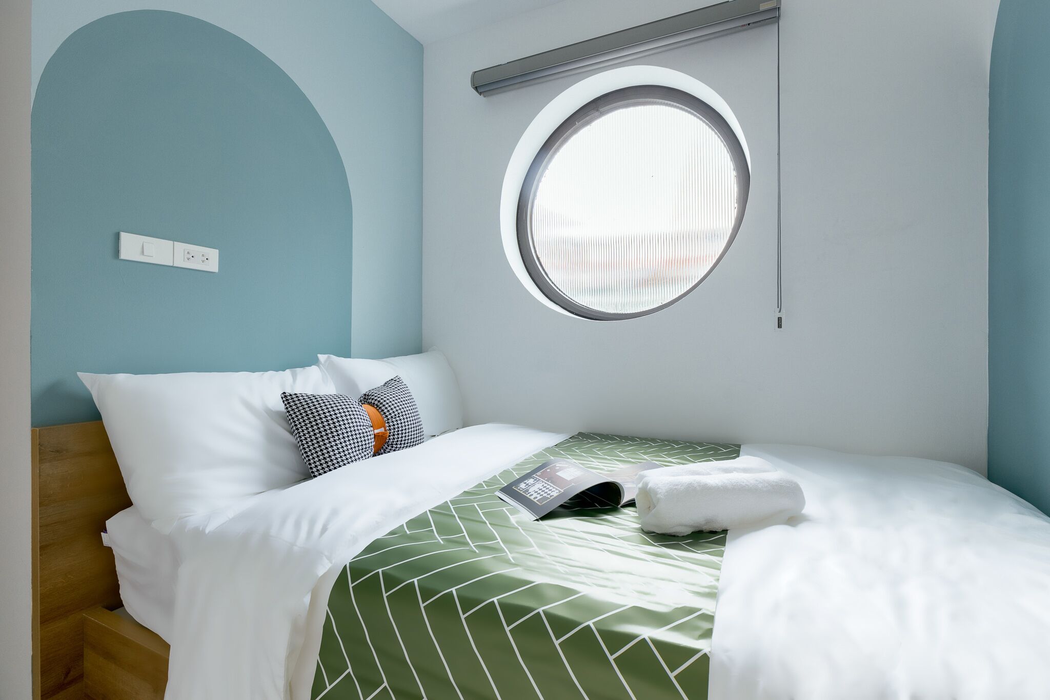 Uma cama bem arrumada com roupa de cama branca e um edredom verde estampado, uma janela redonda com persianas e uma parede azul claro com uma tomada.