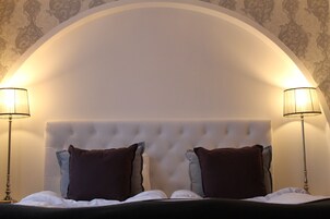 Quarto com cabeceira estofada, duas luminárias de mesa e cama com roupa de cama branca e dois travesseiros.