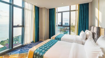 Quarto de hotel com duas camas, janelas amplas com cortinas e vista para a paisagem urbana.