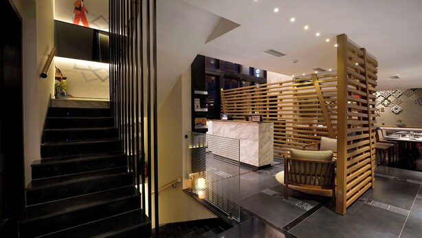 Um lobby de hotel moderno com uma receção em mármore, uma escadaria com grades pretas e uma área de estar com divisórias de madeira.
