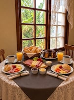 Mesa de café da manhã posta com diversos alimentos, uma janela com cortinas de renda e vista para o verde lá fora.
