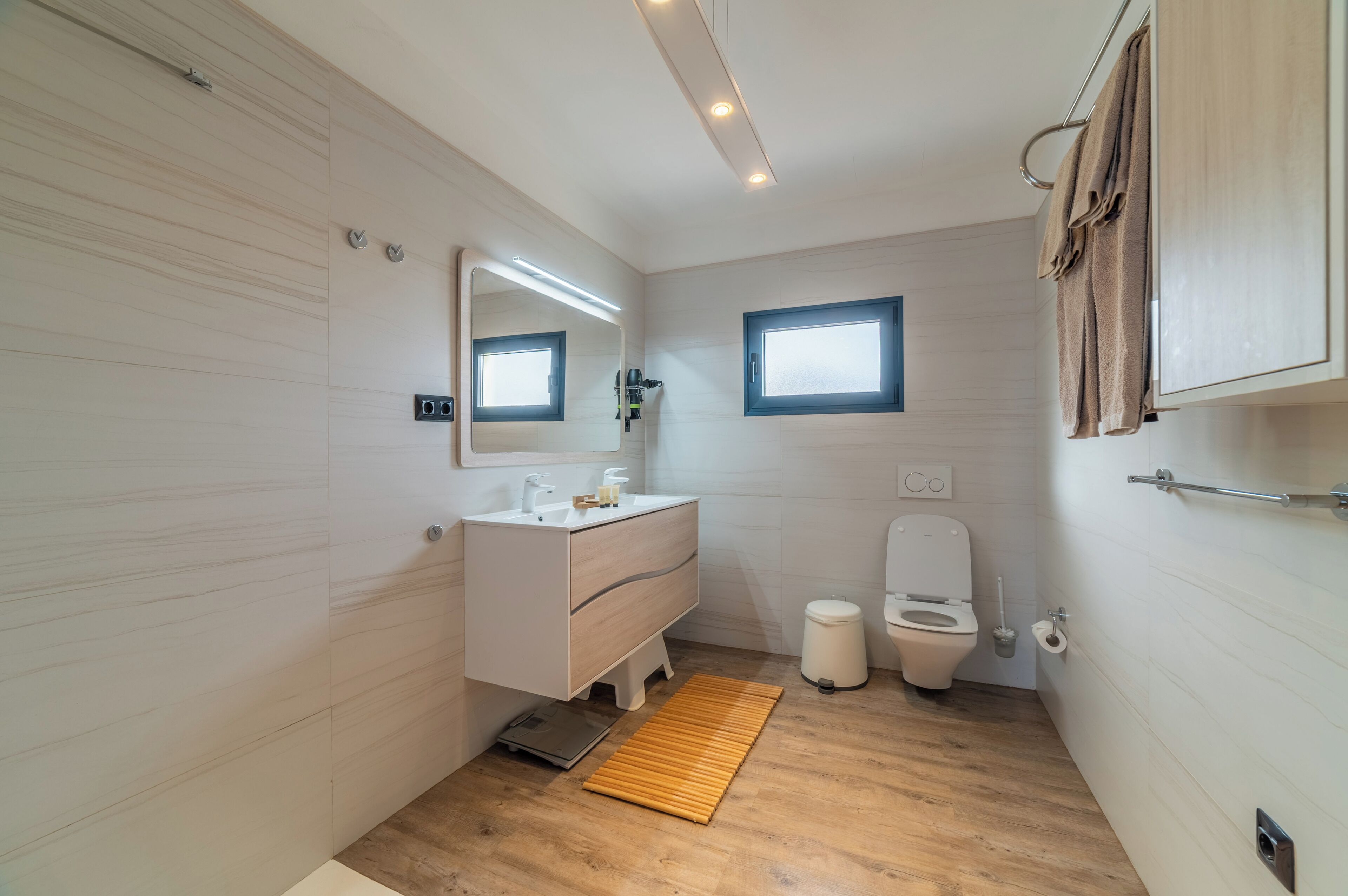 Uma casa de banho moderna com uma toucador branco, um espelho, uma janela, uma sanita e um chão de madeira.