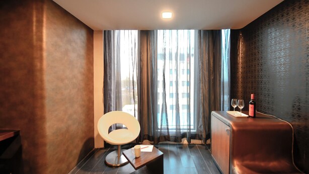 Quarto moderno com sofá de couro, uma poltrona redonda, mesinha com garrafa de vinho e copos, e vista através de cortinas leves.