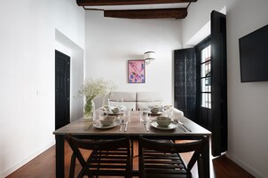 Uma sala de jantar com uma mesa de madeira posta para quatro pessoas, rodeada por cadeiras de madeira escura. Há um vaso com flores brancas na mesa, e um quadro na parede.