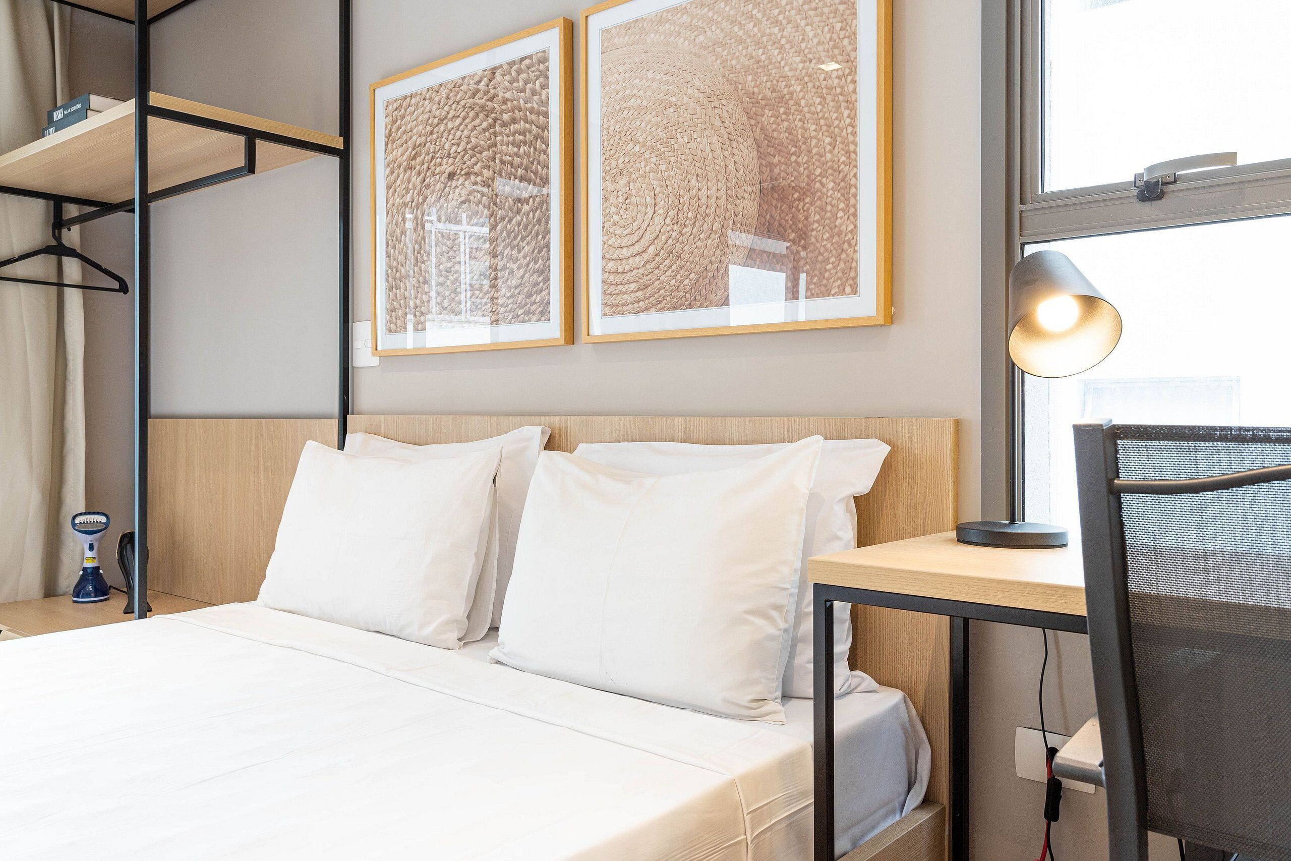 Quarto de hotel com cama, duas almofadas, escrivaninha com abajur, cadeira e dois quadros na parede.