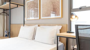 Quarto de hotel com cama, duas almofadas, escrivaninha com abajur, cadeira e dois quadros na parede.