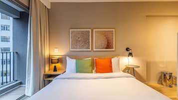 Quarto de hotel com cama, mesa de cabeceira, abajur, duas obras de arte emolduradas, janela com cortinas e varanda.