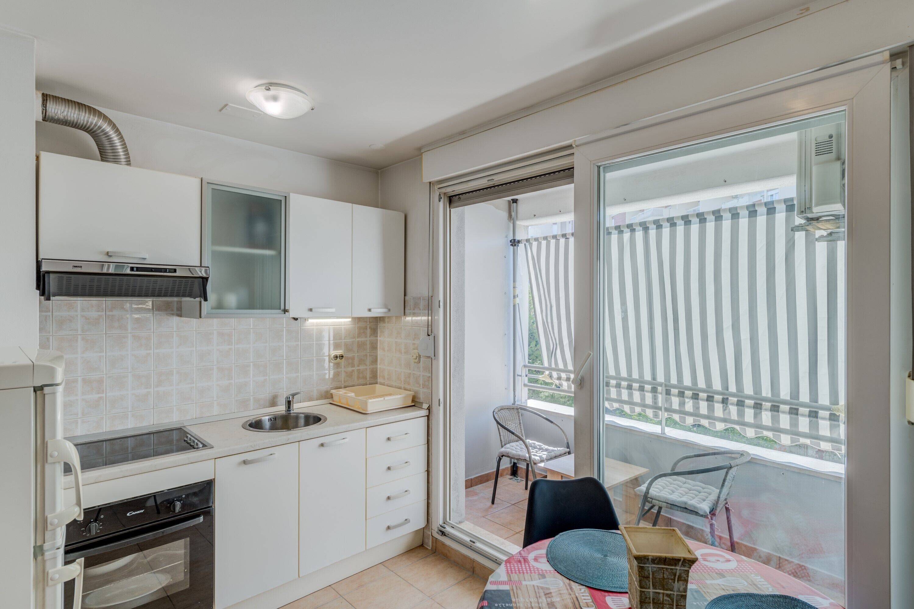 Uma cozinha compacta com armários brancos, forno embutido e pia. É visível uma área de refeições com mesa e cadeiras através de uma porta de vidro que dá para uma varanda com toldo listrado.