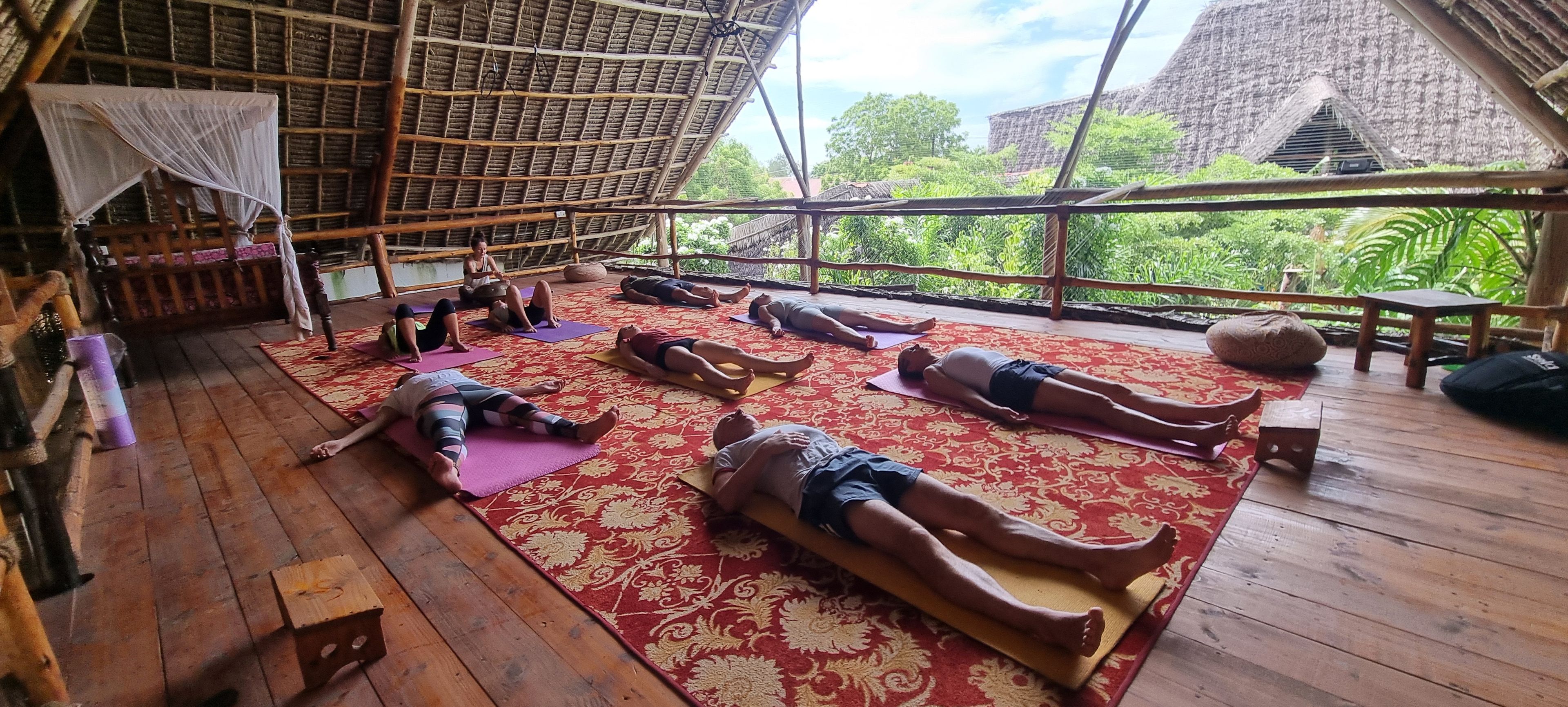 Um grupo de pessoas praticando yoga em tapetes em um quarto com piso de madeira e um tapete vermelho estampado.