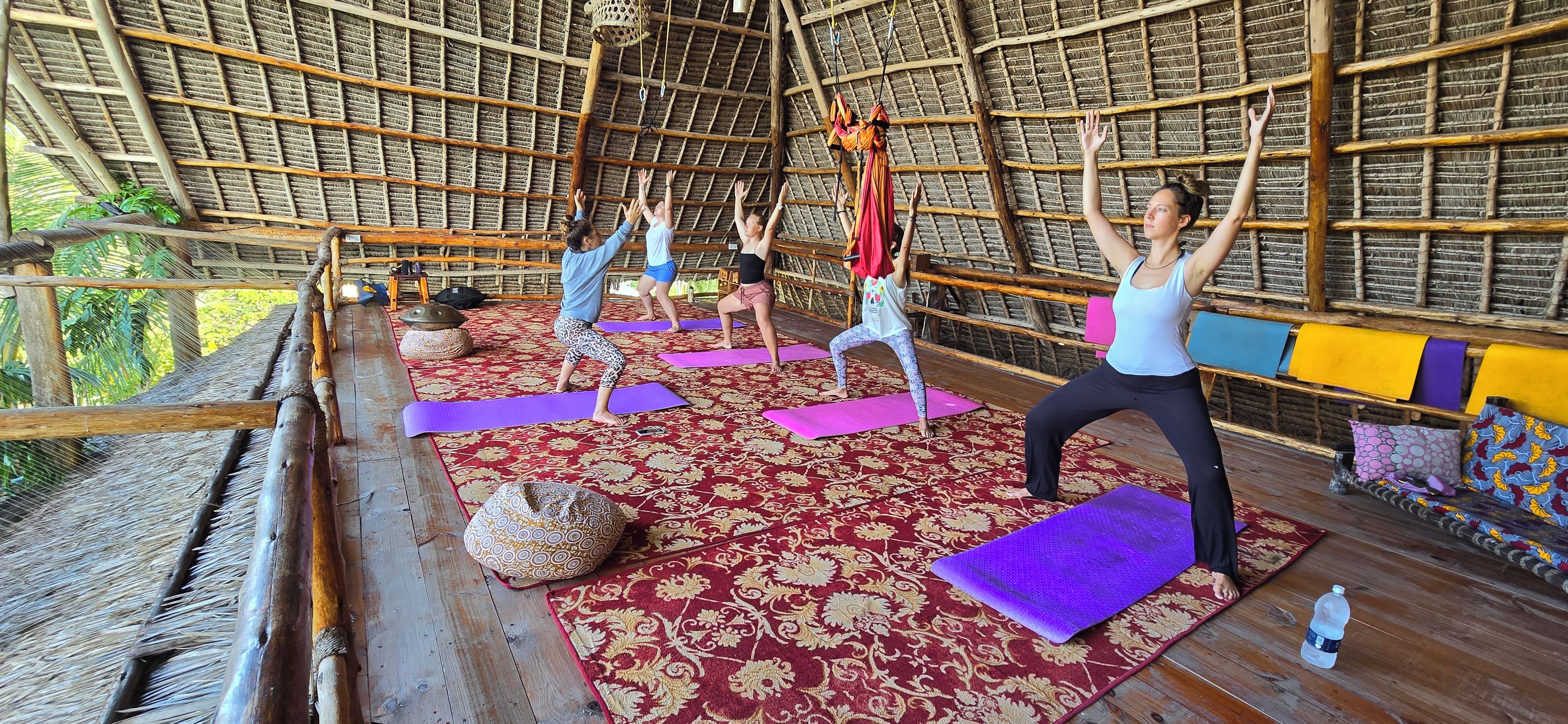 Um grupo de pessoas praticando yoga em tapetes em um espaço com teto de colmo e piso de madeira.