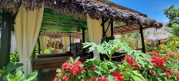 Uma construção com telhado de palha e área coberta para sentar, rodeada por vegetação exuberante e plantas floridas.