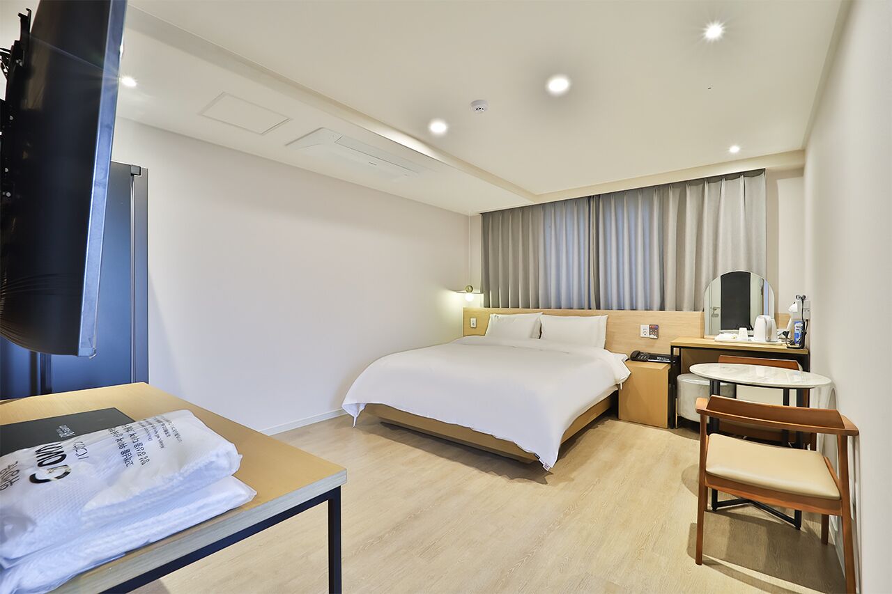 Quarto de hotel com cama, escrivaninha, cadeira, televisão e uma mesinha com toalhas.