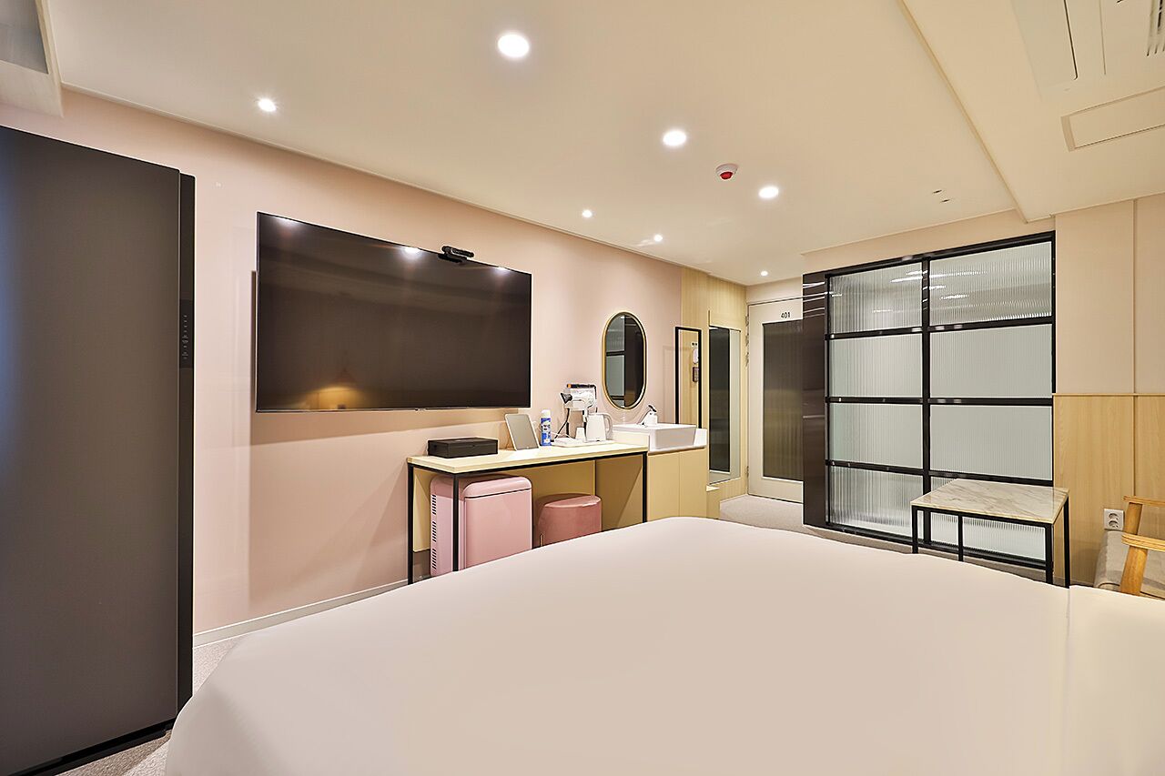Quarto de hotel moderno com uma cama grande, uma escrivaninha com computador, uma toucador com espelho e uma janela ampla.