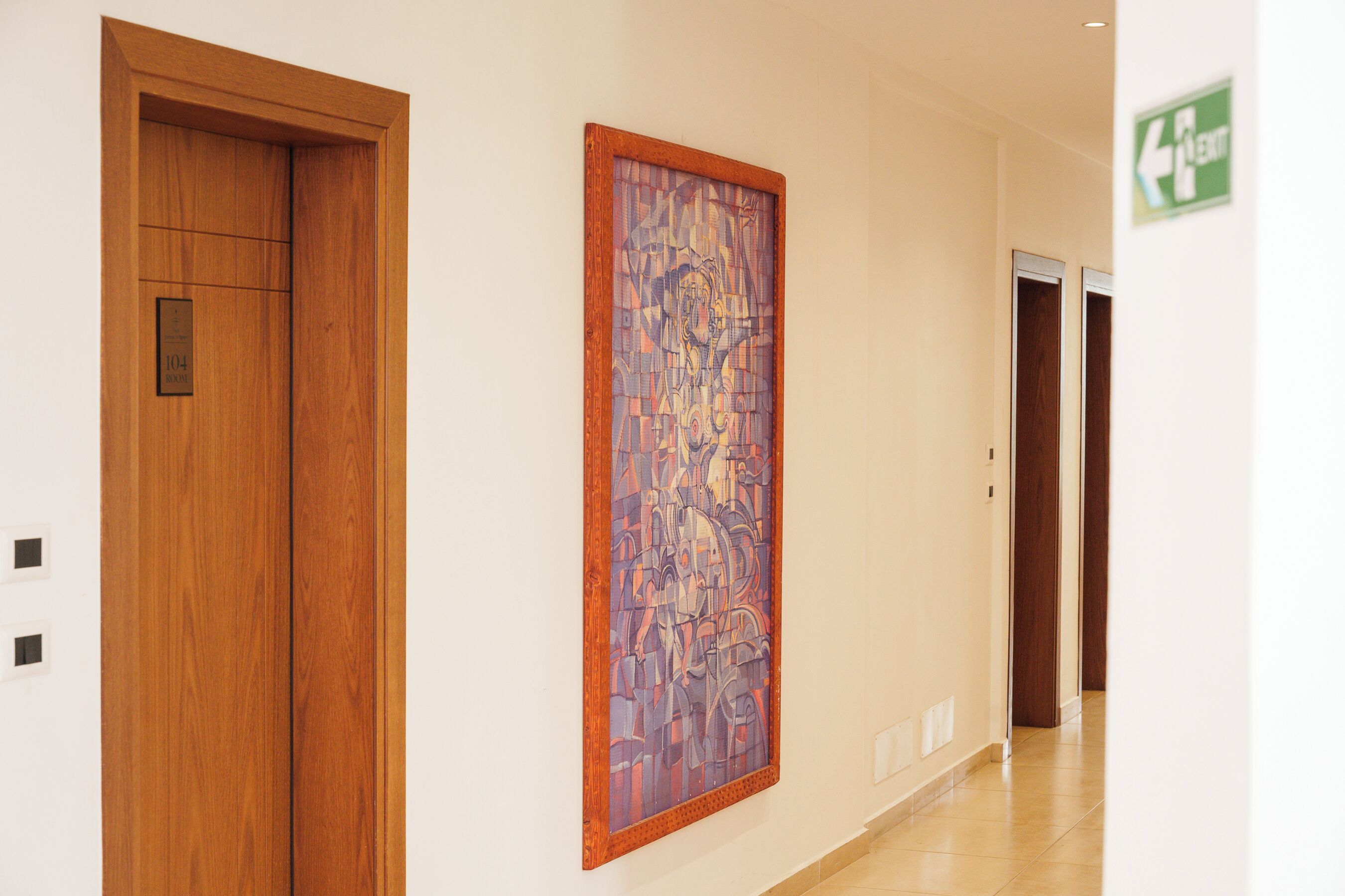 Um corredor com portas de madeira, um quadro abstrato emoldurado e um sinal de saída de emergência verde.