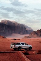 Uma caminhonete prateada estacionada no deserto, com formações rochosas ao fundo.