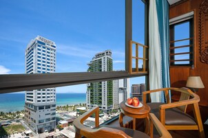 Uma varanda com vista para o oceano e edifícios altos, uma cadeira de madeira, uma mesa pequena com frutas e um abajur.