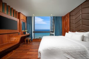 Quarto de hotel com uma cama grande, um banco de madeira, televisão de tela plana e vista para o oceano.