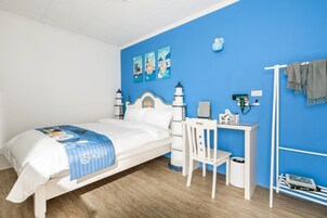Quarto com cama, escrivaninha e cadeira. Há decorações nas paredes e uma mesinha com abajur.