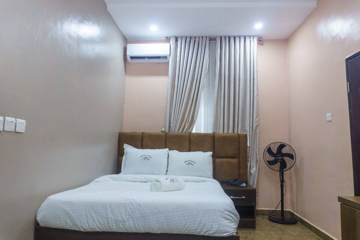 Quarto com cama, mesa de cabeceira, ventilador e janela com cortinas.