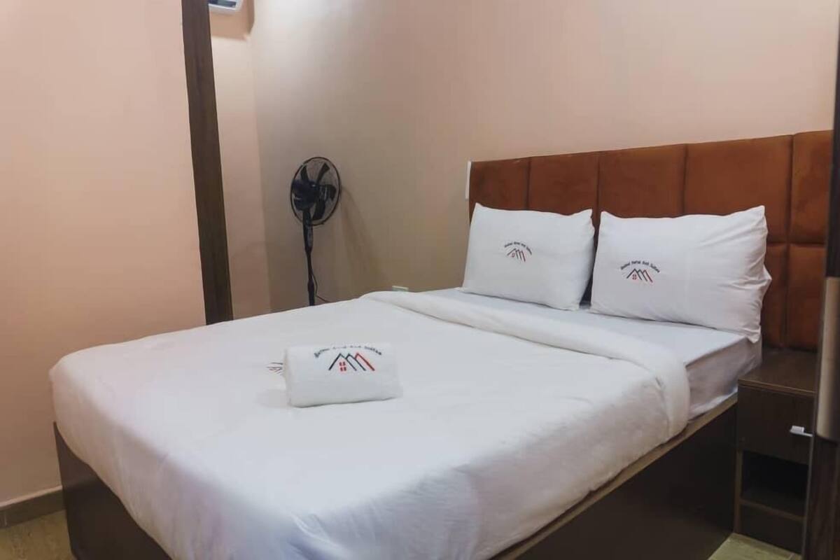 Cama bem arrumada com lençóis e almofadas brancos, cabeceira marrom e ventilador fixado na parede.