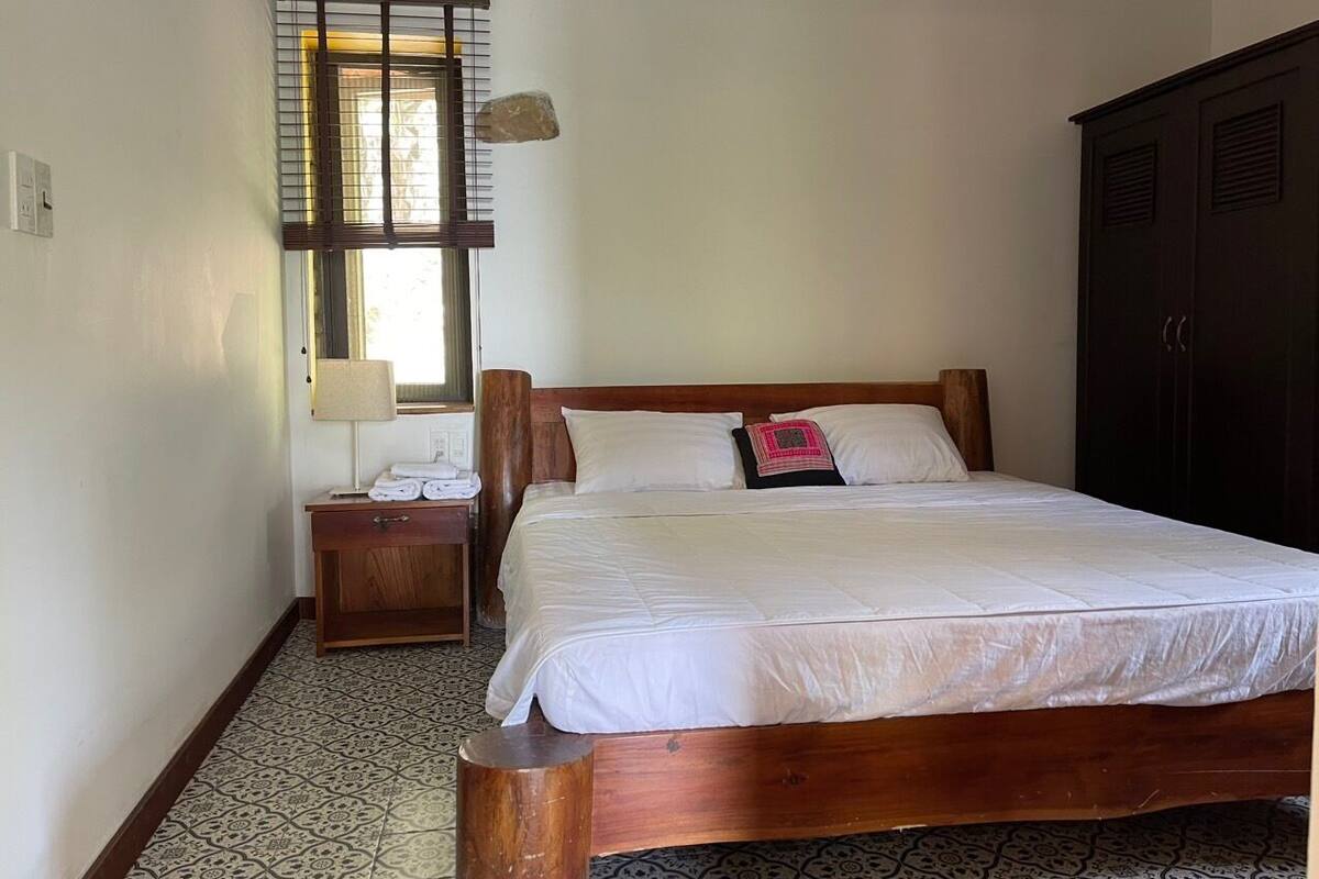 Uma cama bem arrumada com cabeceira de madeira, mesa de cabeceira com abajur e guarda-roupa.